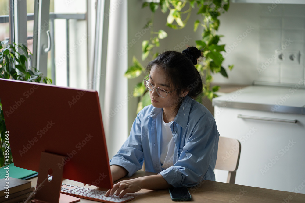 Focused Asian woman IT developer wearing glasses writing code while working remotely from home ...