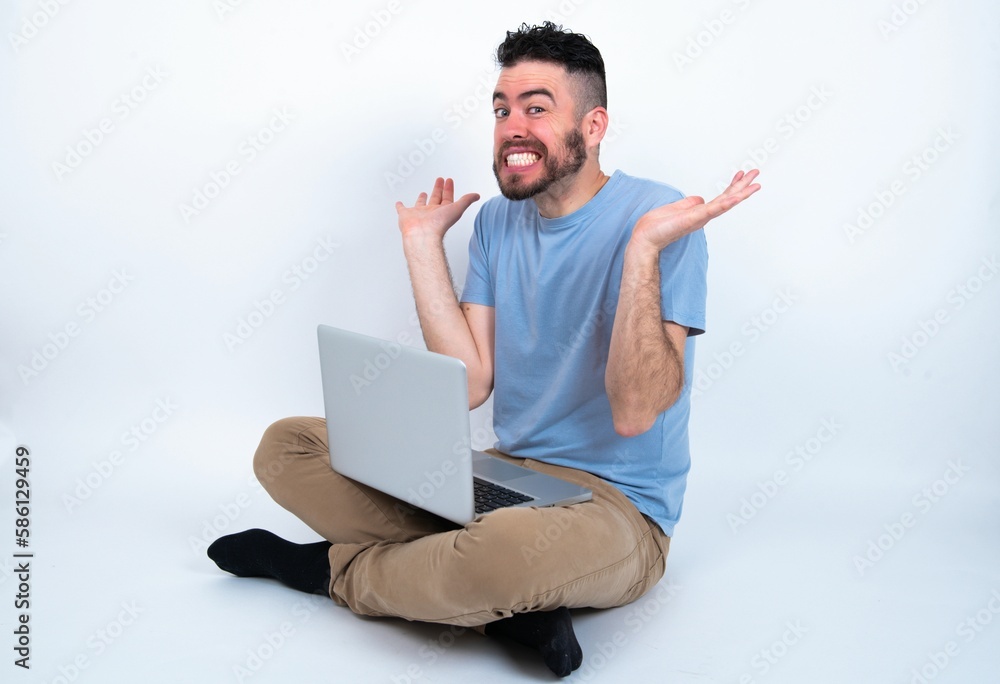 Clueless Young caucasian man with laptop sitting over white studio ...