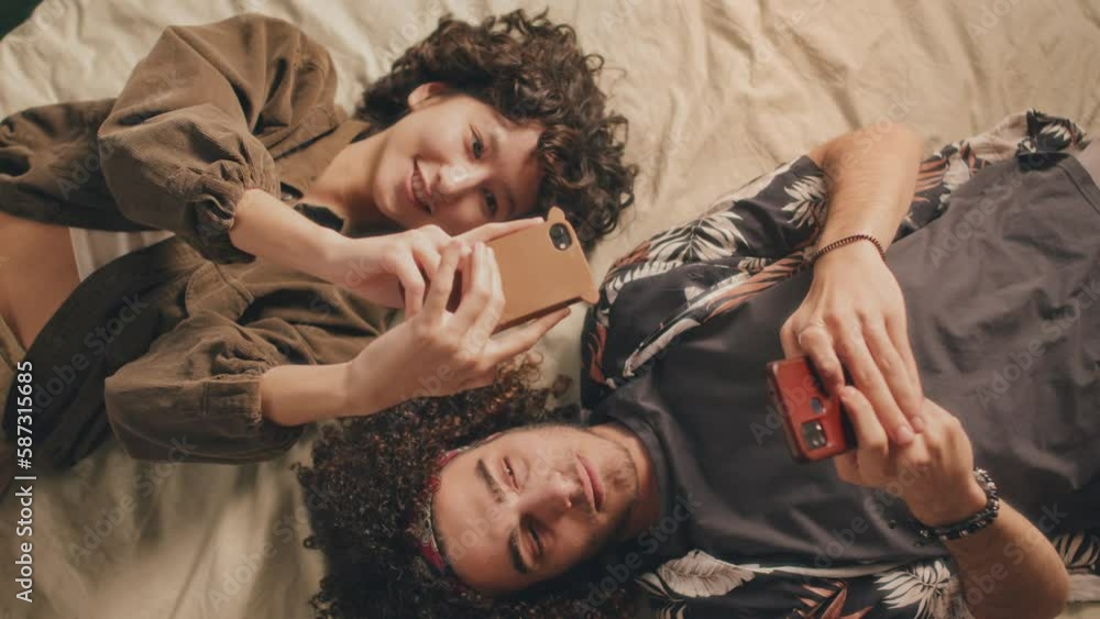 Teenage couple lying in bed with phones, head to head, looking and ...