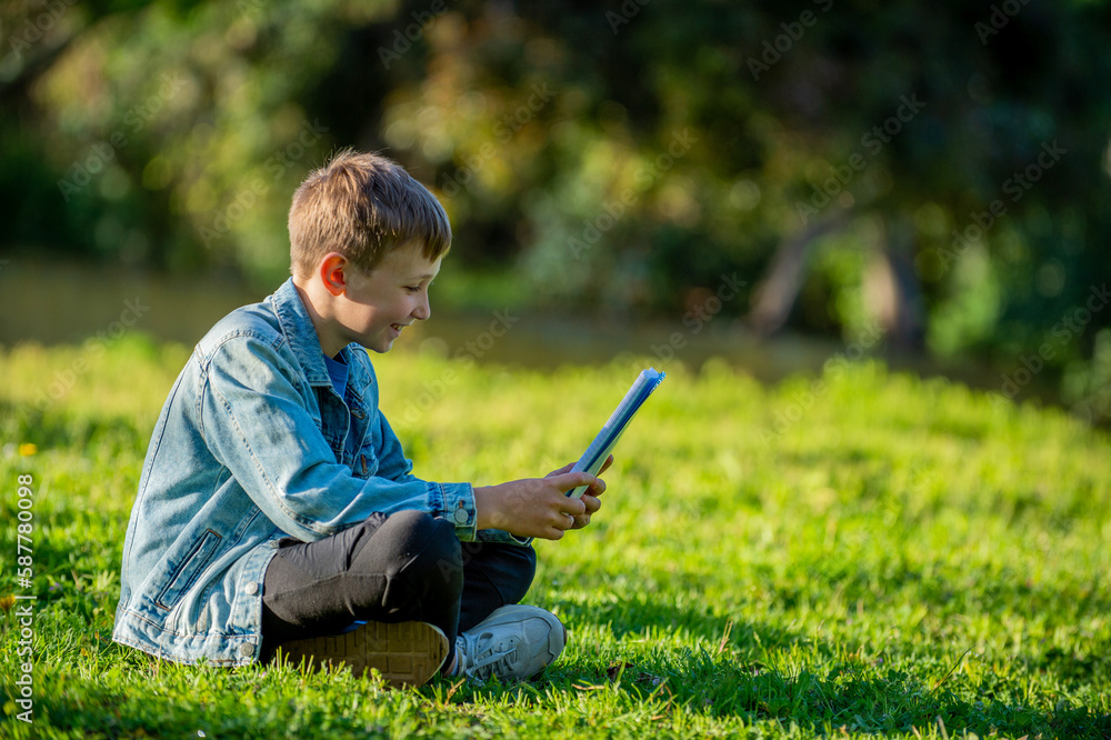 Happy scholar with a textbook in his hands is sitting on a green lawn ...