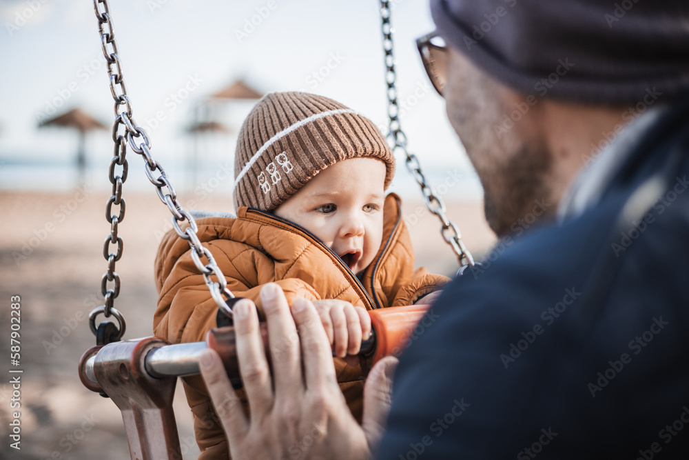 Father pushing hir cheerful infant baby boy child on a swing on sandy ...