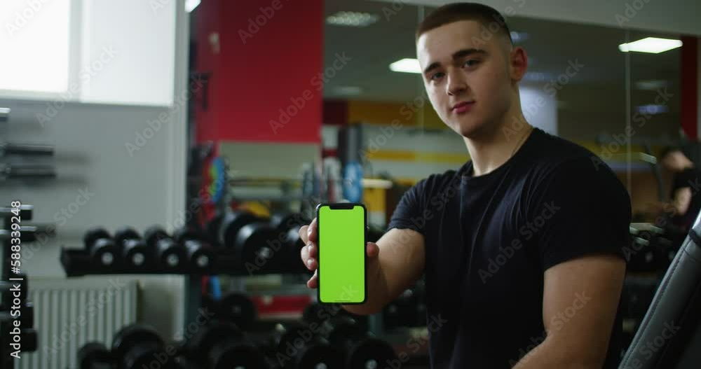 Man athletic build in gym shows smartphone with green screen to camera ...