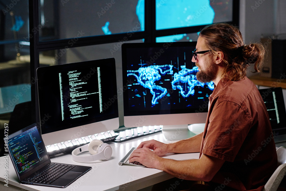 Young male programmer in eyeglasses looking at coded data on computer screen and typing while ...