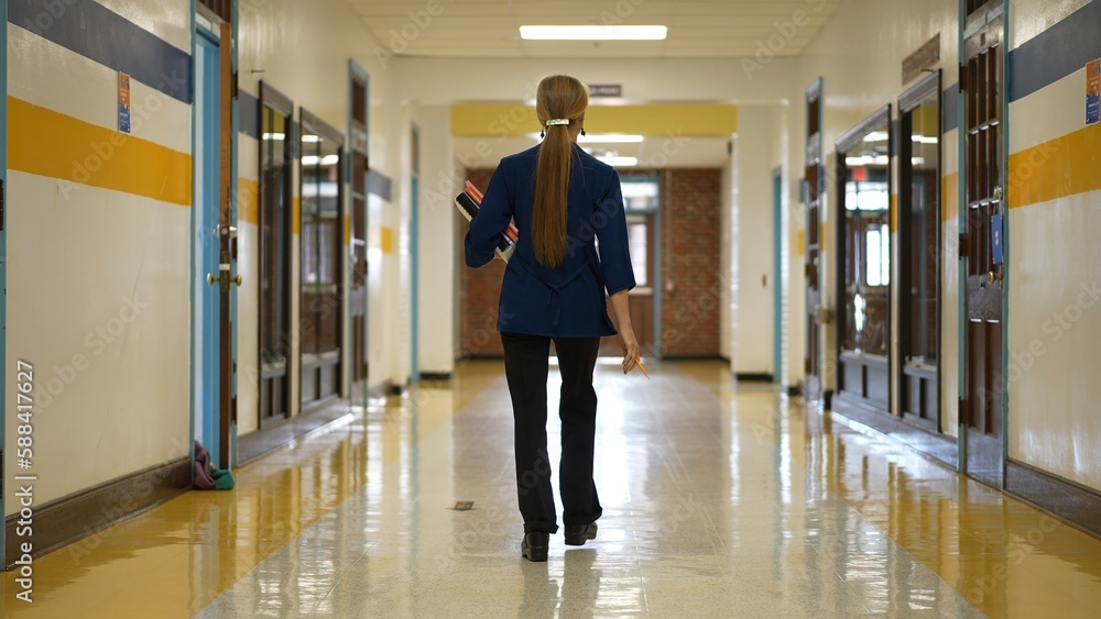 Rear view of teacher walking down a hallway in an empty school holding ...