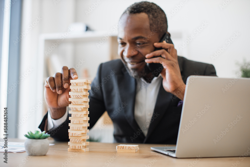 Happy multicultural man in business suit playing jenga while talking on ...