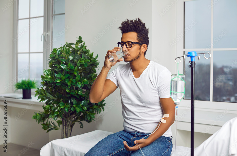 Young African American man drinking glass of water while sitting on medical bed at clinic and ...