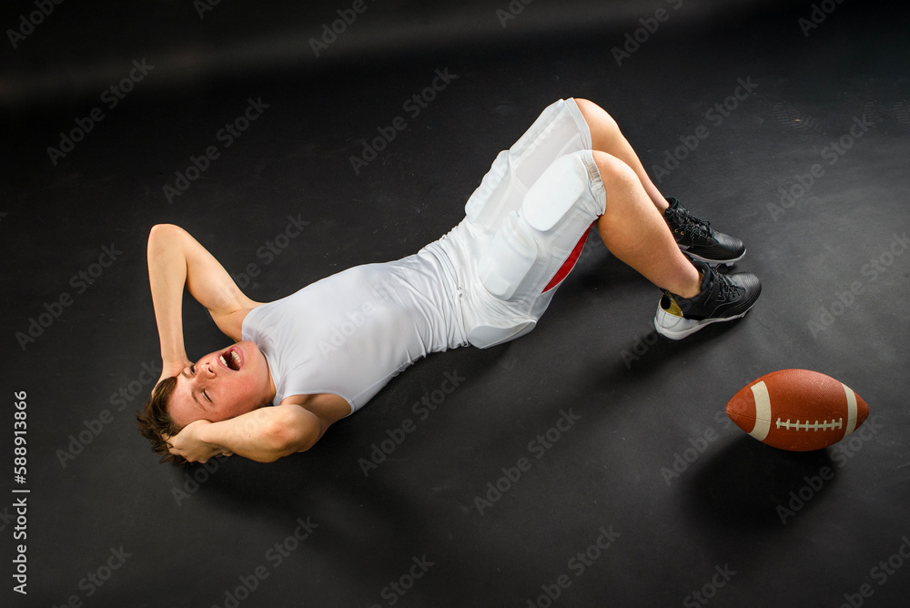 Young preteen football boy in football uniform laying injured on studio ...