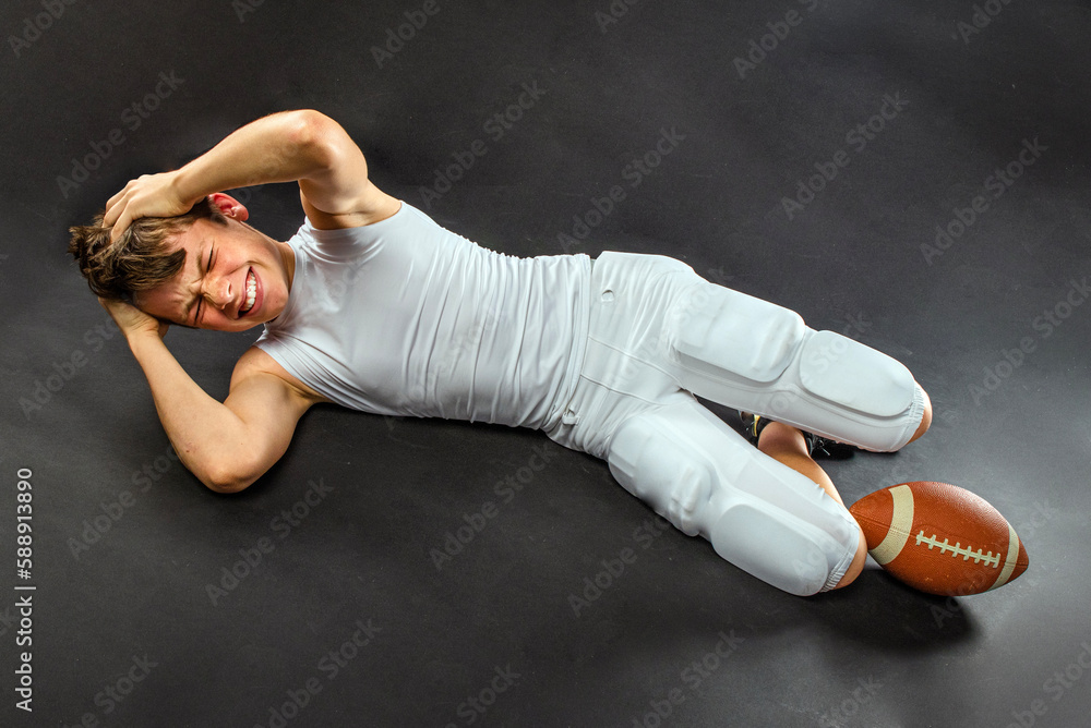 Young preteen football boy in football uniform laying injured on studio ...