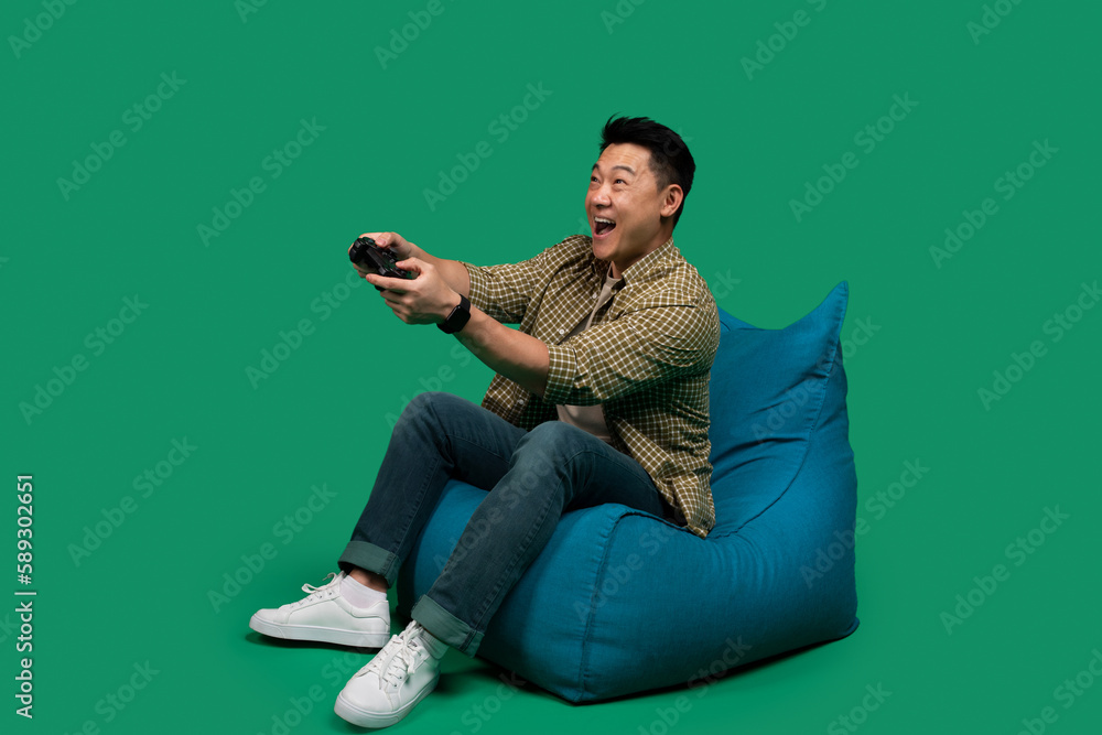 Joyful asian man playing video games with joystick, sitting on beanbag ...
