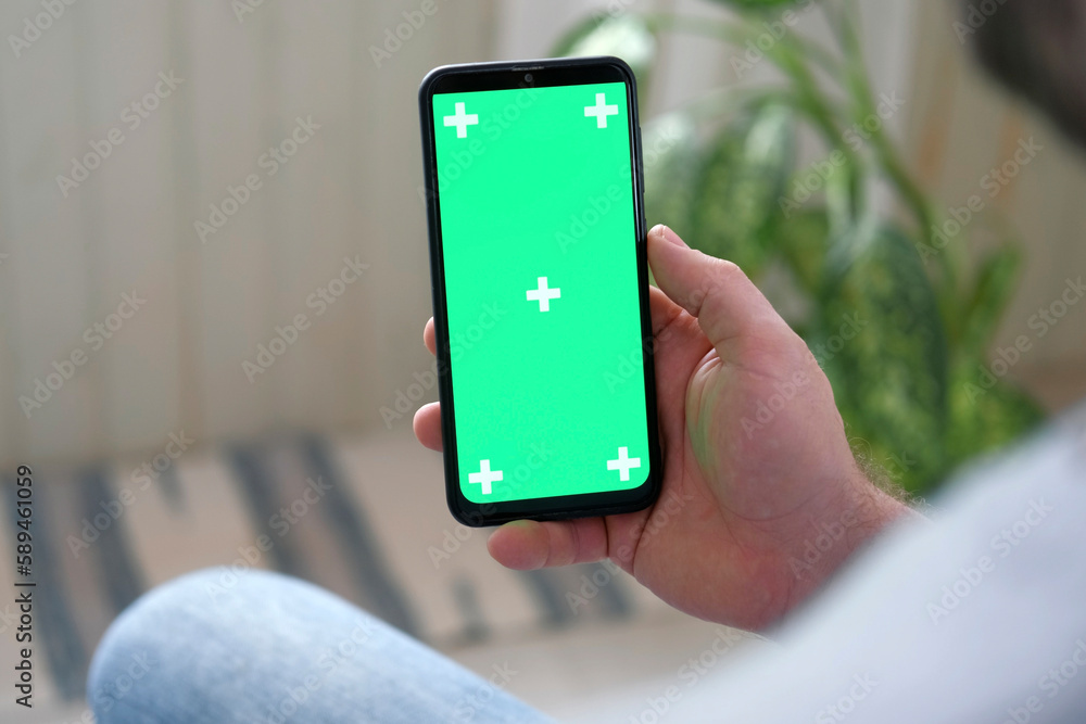 Young man sitting at home holding smartphone green mock-up screen in ...