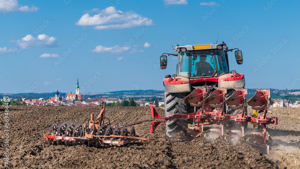 Red tractor with plow and disc harrow in brown arable soil under summer ...