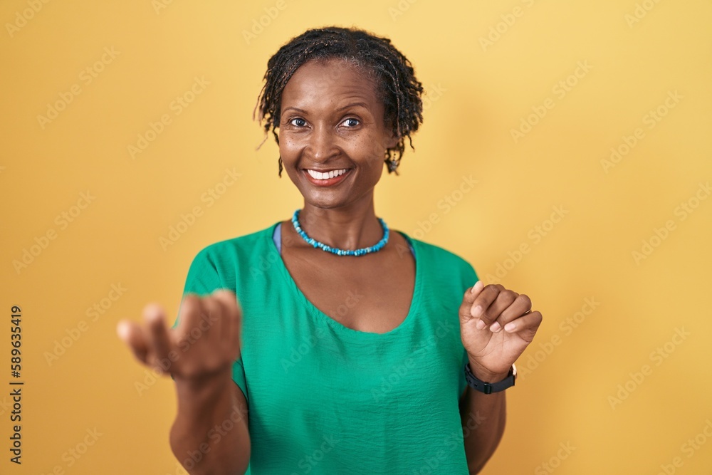 African woman with dreadlocks standing over yellow background beckoning ...