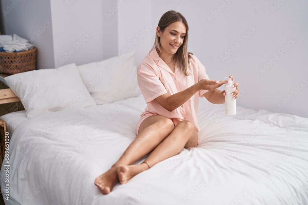 Young caucasian woman applying skin lotion treatment sitting on bed at ...