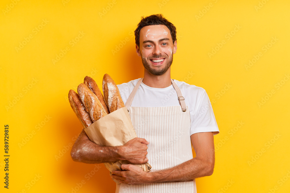 Cute young happy baker holding paper bag with fresh French bread in his ...