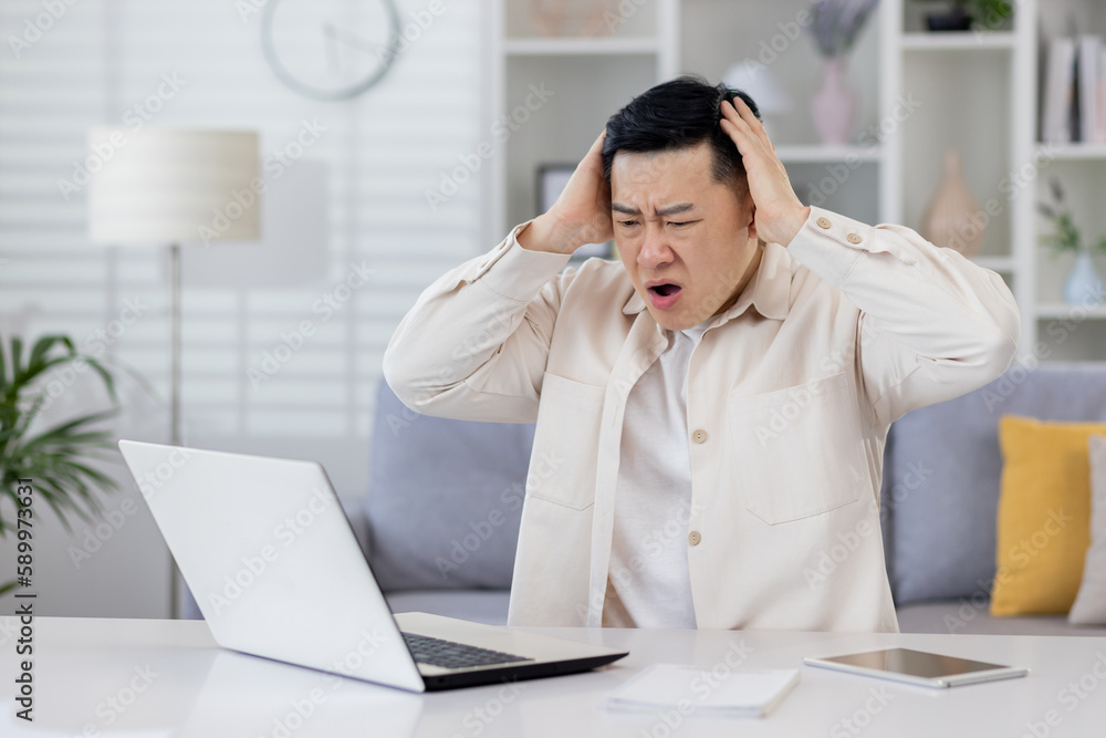 Angry man at home working with laptop, frustrated asian man looking at ...
