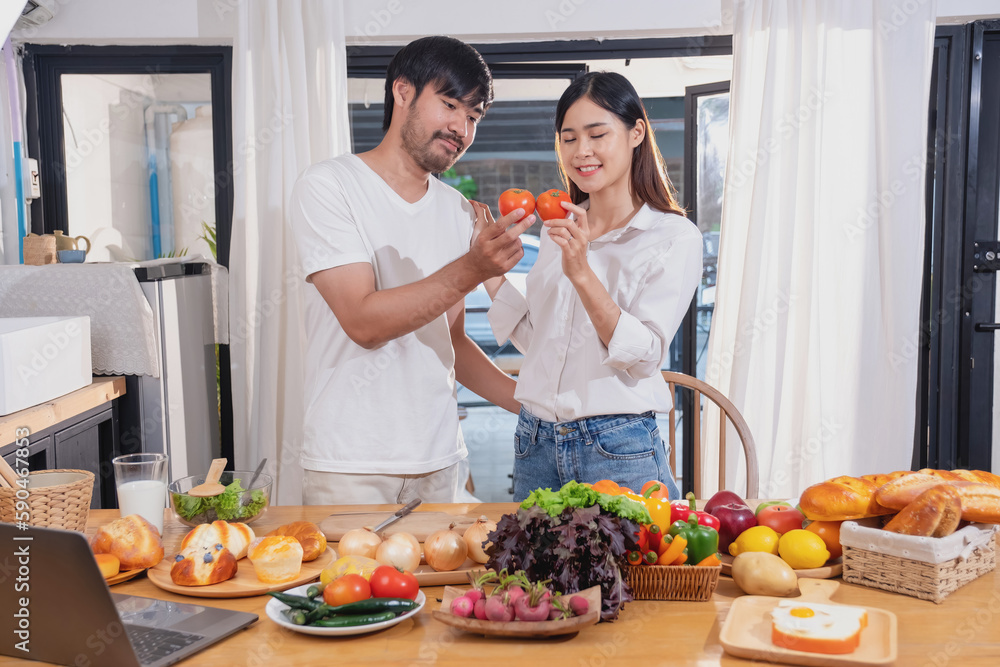 Young Asian couple cooking with fruits and vegetables and using laptop ...