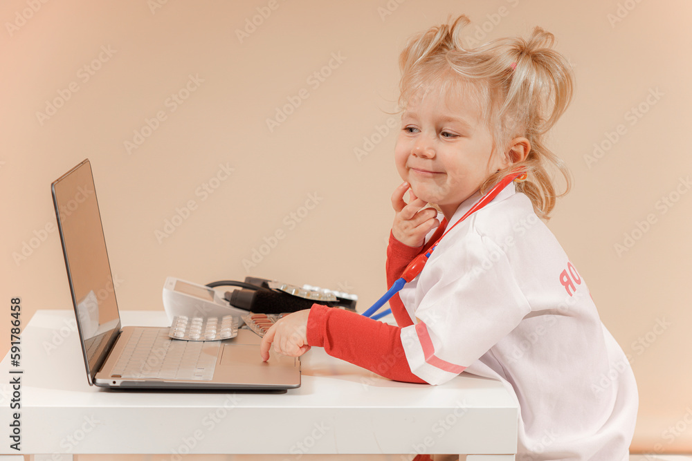 Little girl doctor sits near the laptop and looks ahead. On the table ...