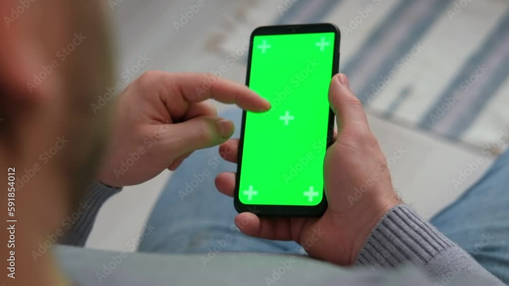 Young man sitting at home holding smartphone green mock-up screen in hand. Male person using ...