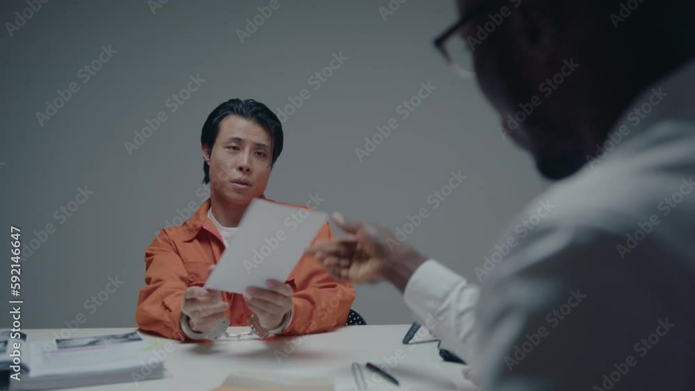 Asian criminal in handcuffs and jail uniform looking at evidence photos ...