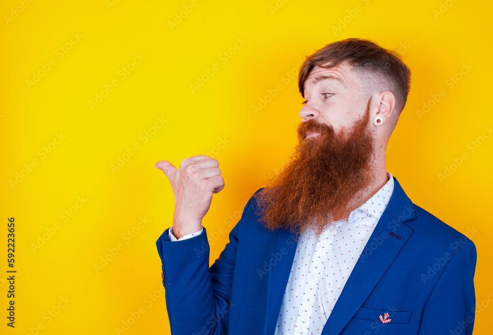 Charming red haired man wearing blue suit over yellow studio background looking at copy space ...