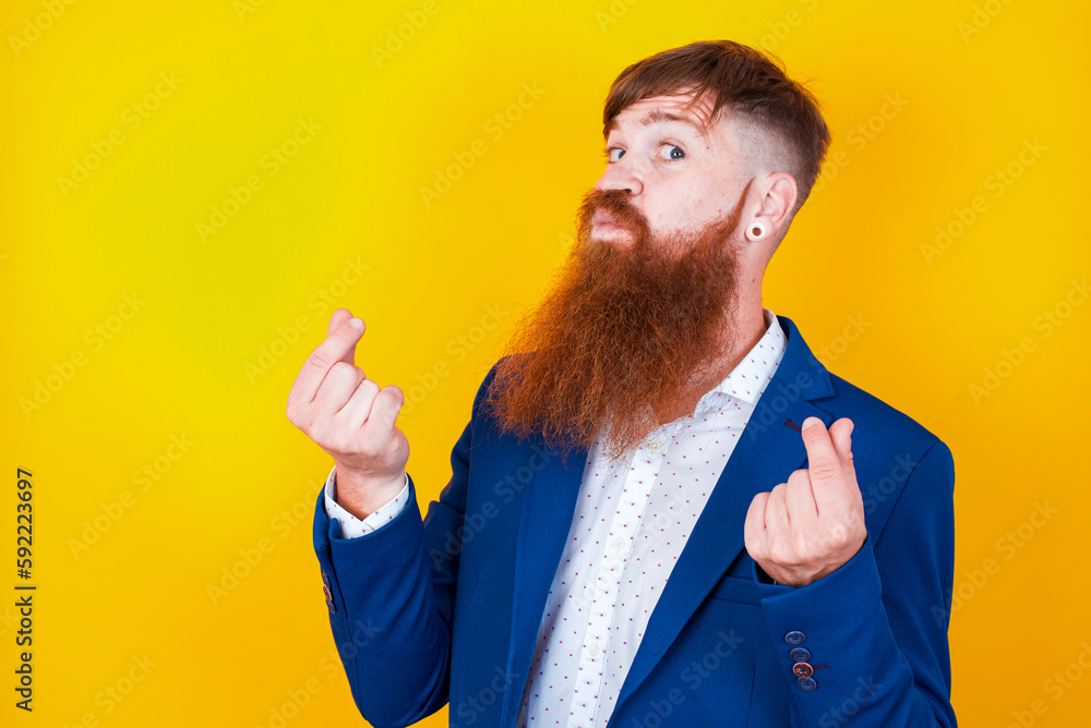 red haired man wearing blue suit over yellow studio background doing money gesture with hands ...