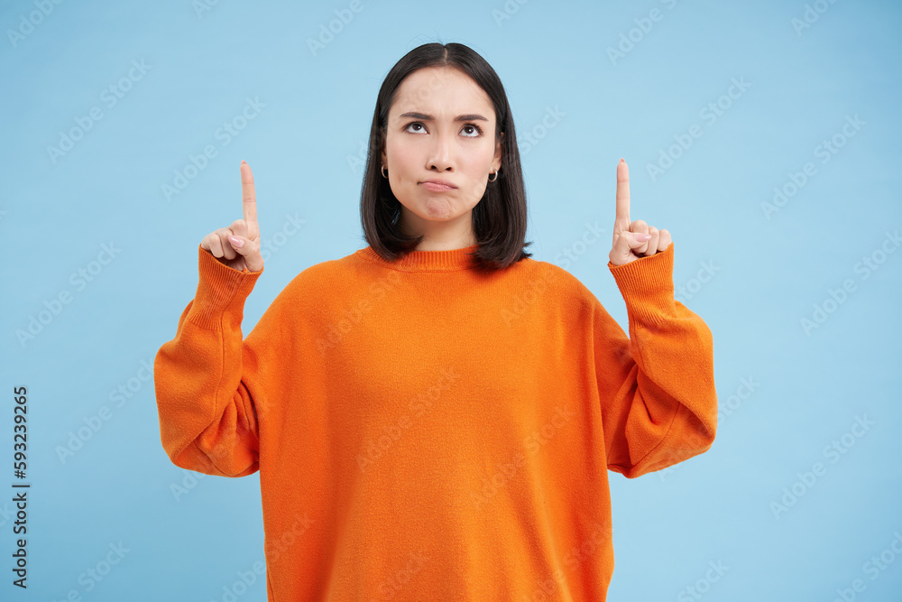 Perplexed asian girl, looks and points up with frustrated, thinking ...
