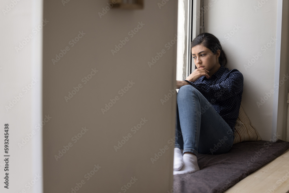 Frustrated pensive Indian woman sits on windowsill and looks out window, suffer from apathy ...