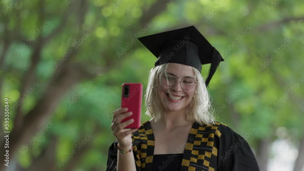 White Female University Graduate wearing cap and gown takes selfie with ...