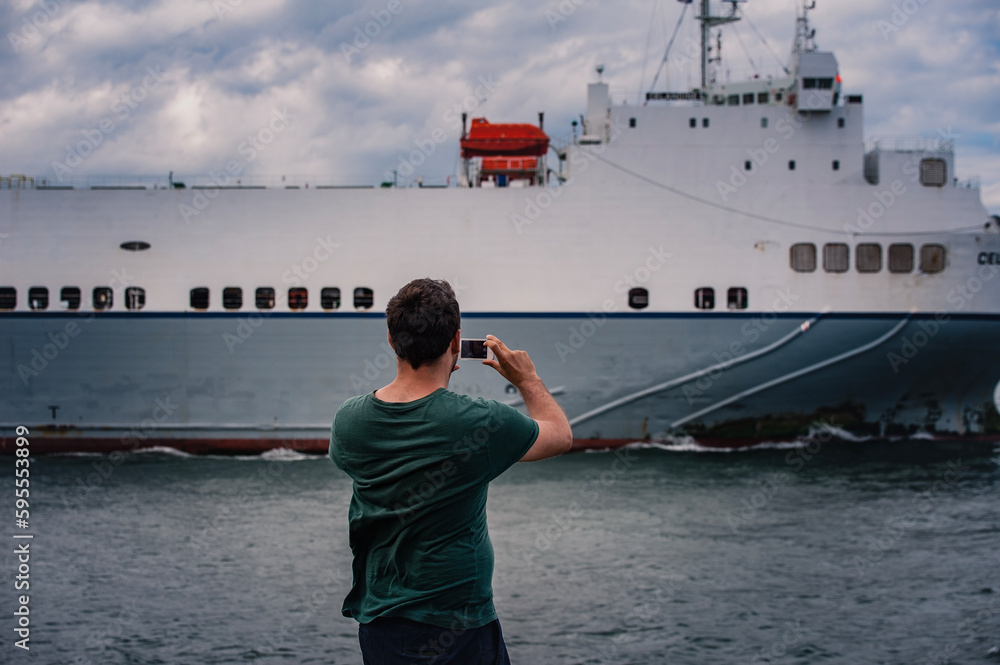 Back view of a person taking a photo of a big passing vessel blurred ...