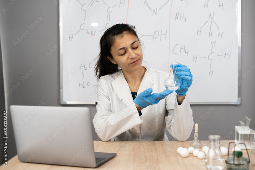 Smiling female chemistry teacher wearing white coat with laptop ...