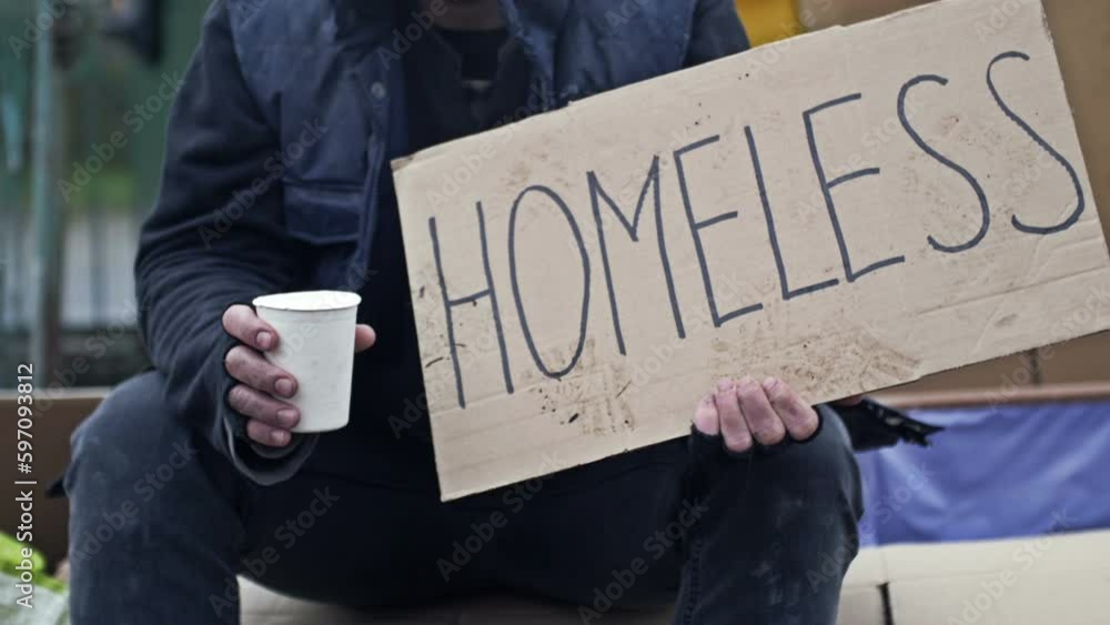 An unkempt-looking man sits with a begging cup and holds a handwritten ...