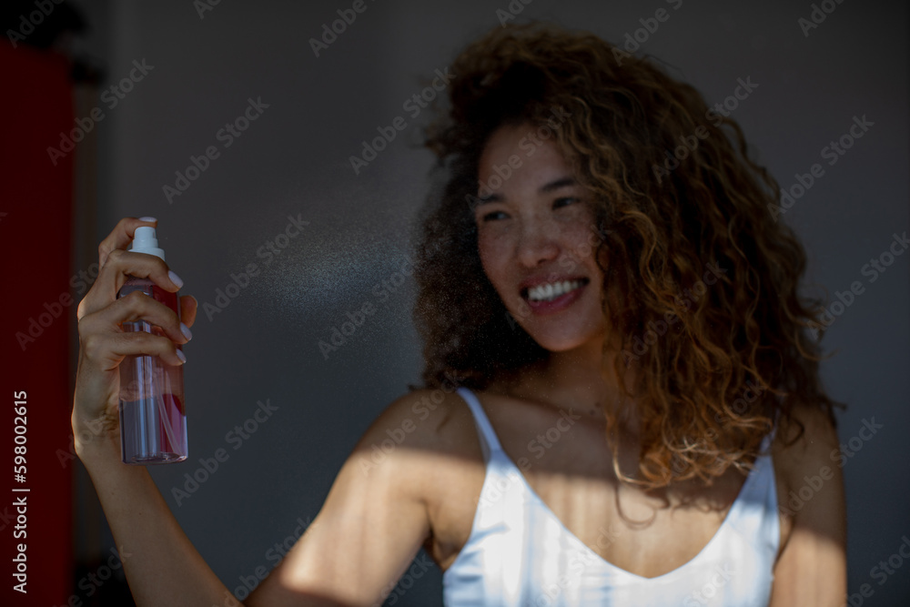 Smiling girl with lush curly hair and freckles on her face is spraying ...