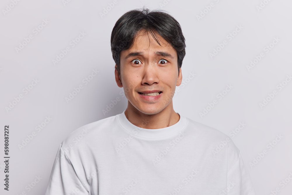 Shocked worried Japanese man has anxious expression clenches teeth stares embarrassed at camera ...