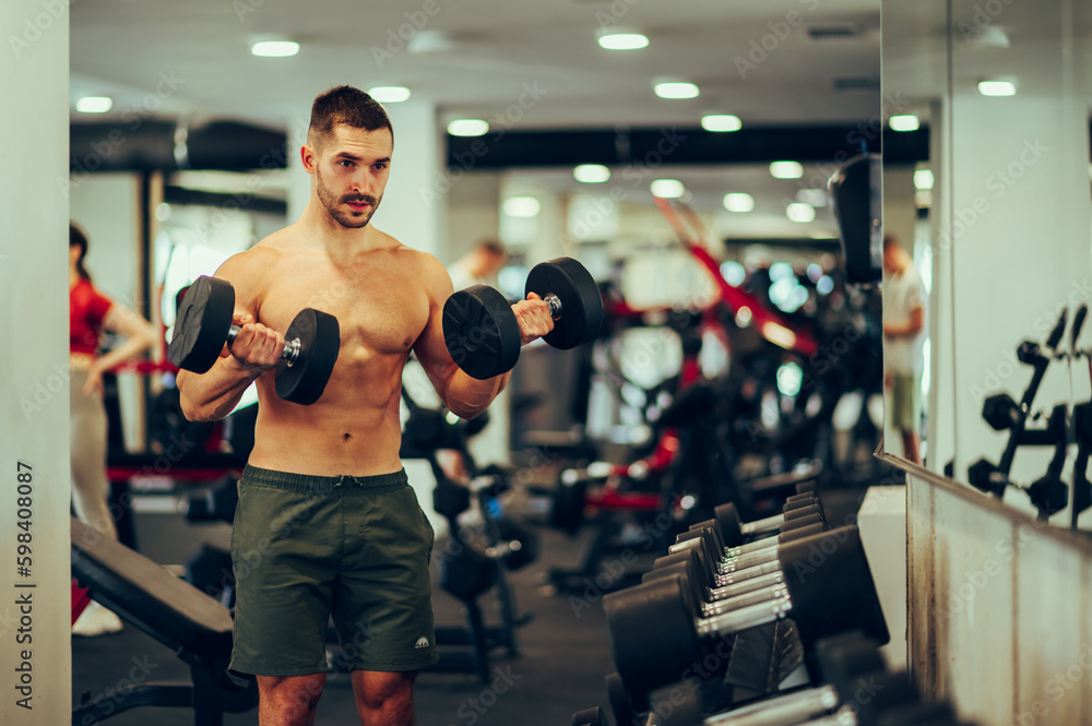 A young fit shirtless man is lifting dumbbells in a gym and working out ...