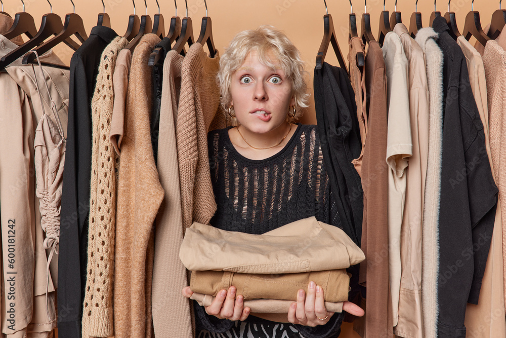 Worried blonde woman bites lips stares embarrassed holds folded clothes ...