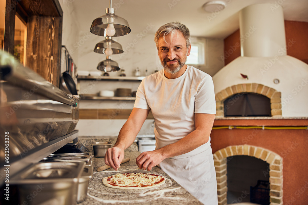 Portrait of a smiling male pizza master, putting a cheese on top of the ...