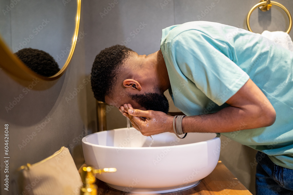Tired sleepy African American guy washing face with water in morning, black man standing in ...