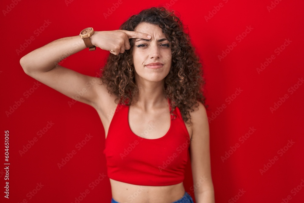 Hispanic woman with curly hair standing over red background pointing ...