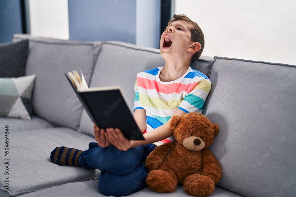 Young caucasian kid sitting on the sofa reading a book angry and mad ...