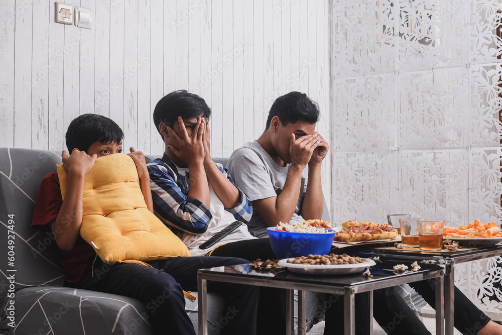 Young scared Asian siblings sitting on sofa and frightenedly watching ...