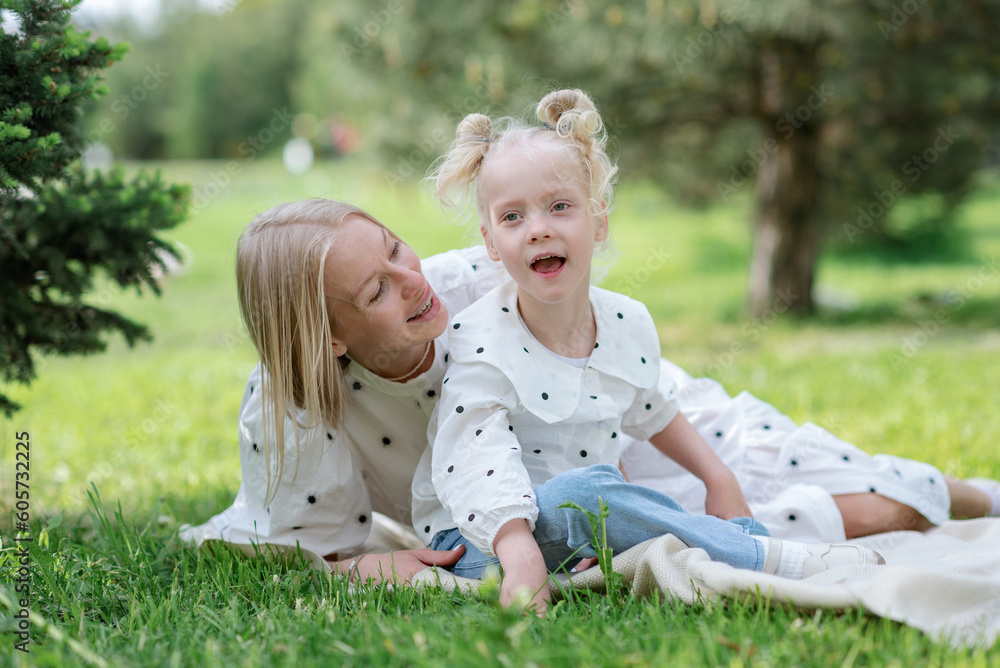 Blond young mother with disabled daughter sitting ot the green grass in ...