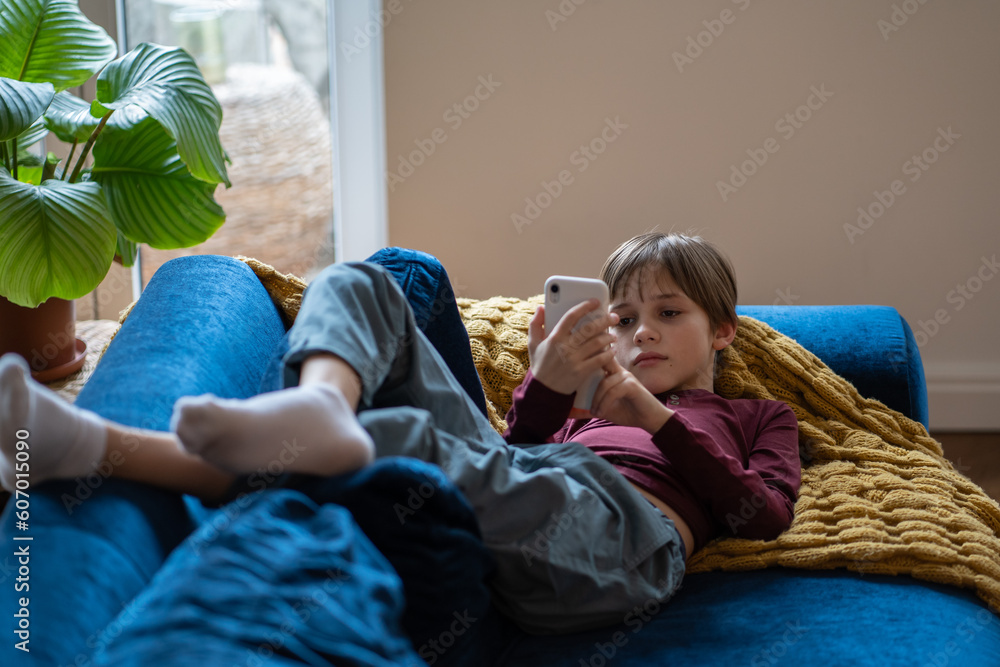 Depressed sad little kid schoolboy lying on sofa looking on mobile ...