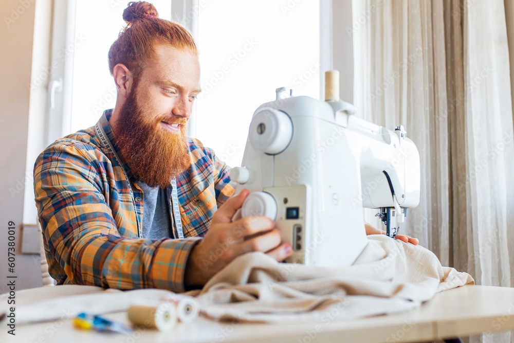 handsome redhaired man with long beard sews at a sewing machine at home ...