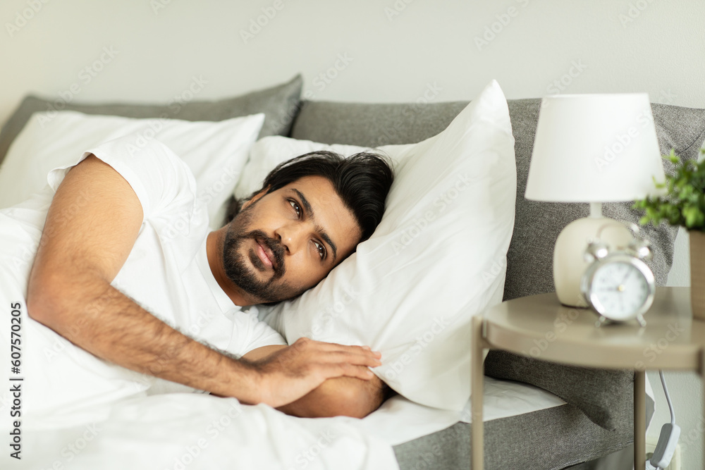 Sad serious middle eastern man woke up, lies on bed, looks at alarm ...