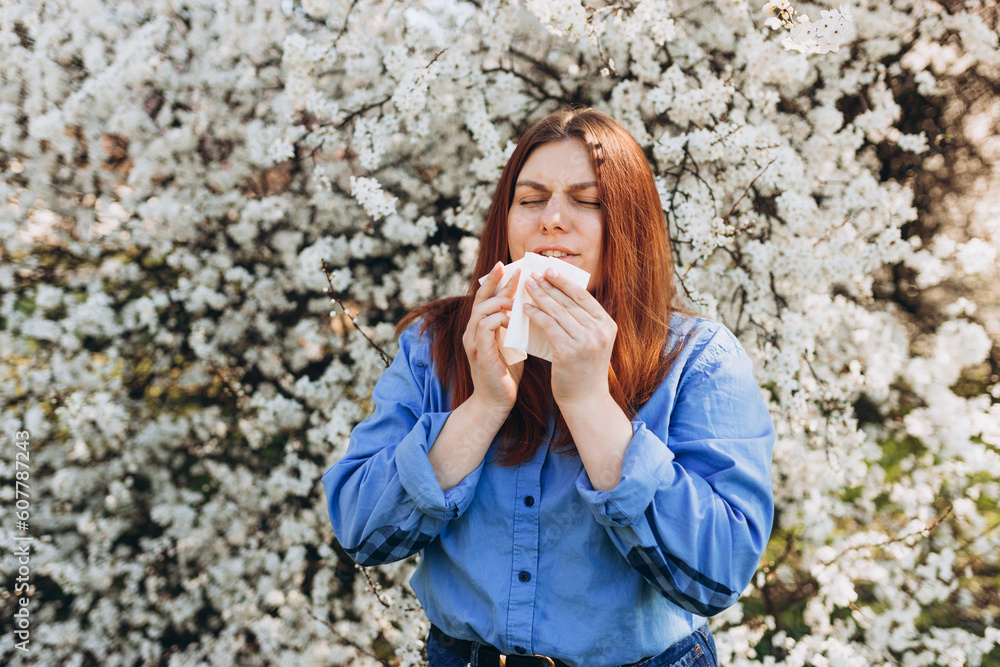 Sneezing young redhead woman with nose wiper among blooming trees in ...
