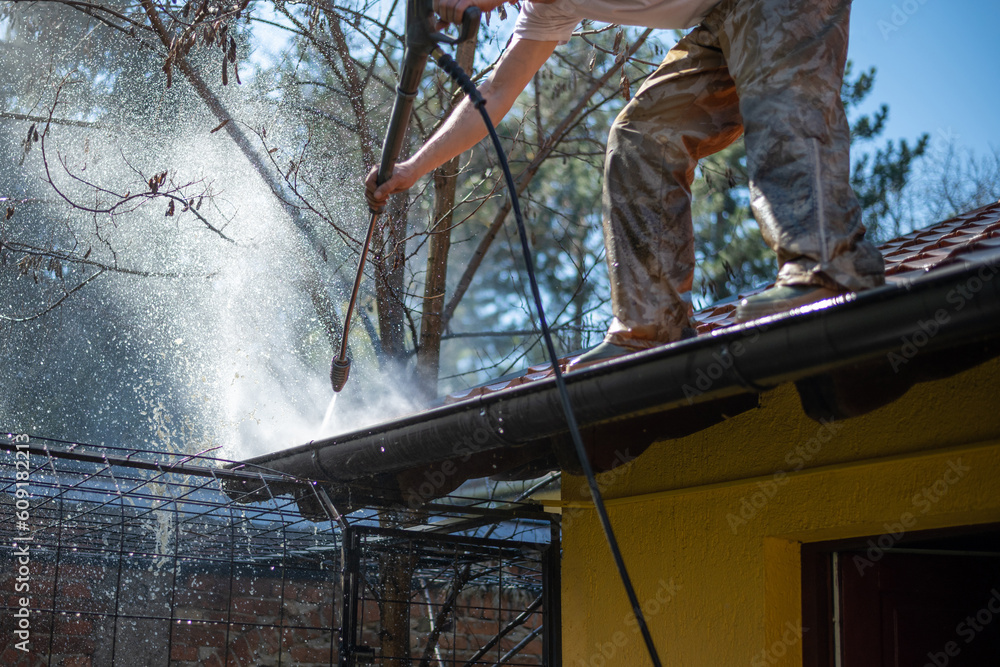 Worker standing on the roof and cleaning rain gutter with high pressure ...