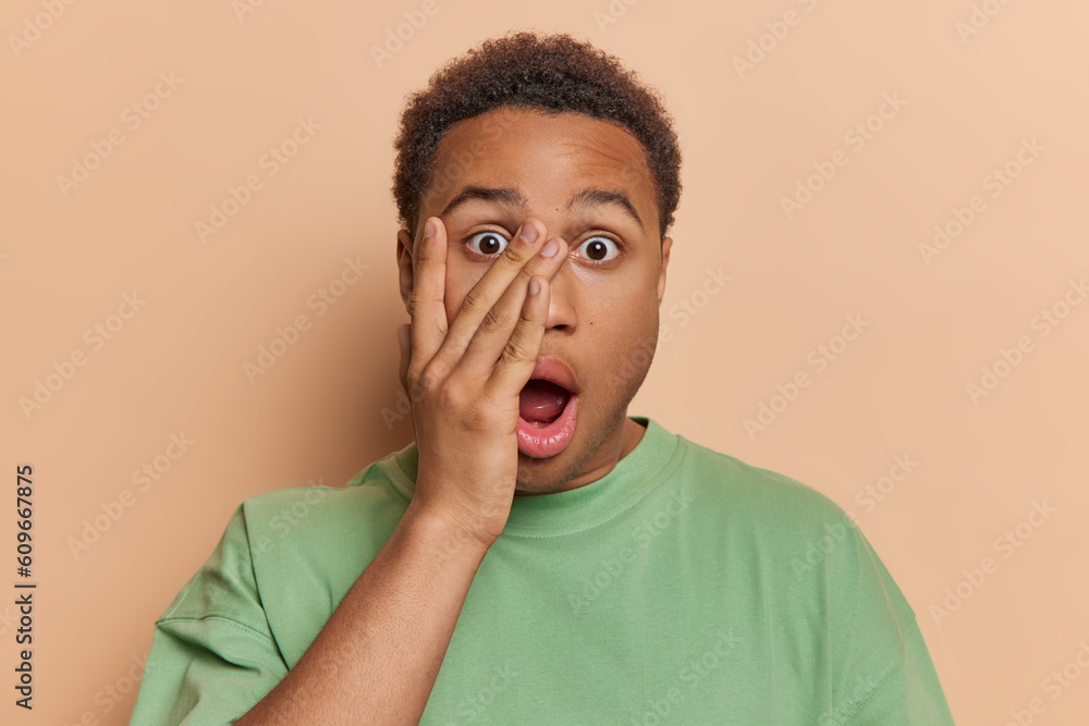 Frightened dark skinned man covers face with hand keeps mouth widely ...