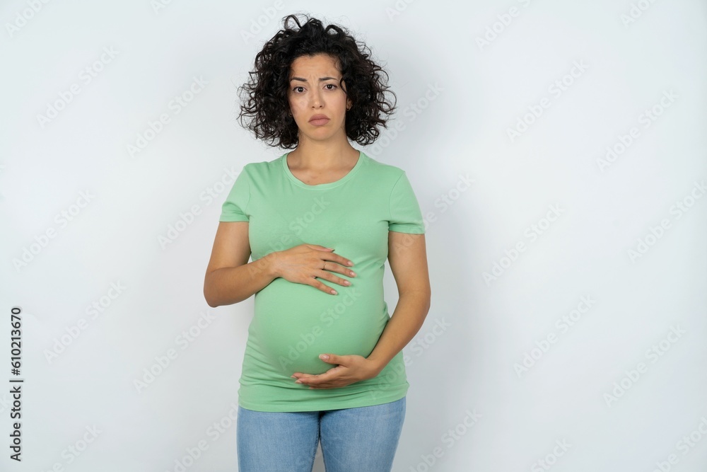 Displeased young pregnant woman wearing green t-shirt over white background frowns face feels ...