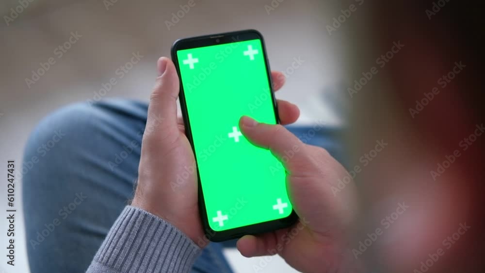 Young man sitting at home holding smartphone green mock-up screen in hand. Male person using ...