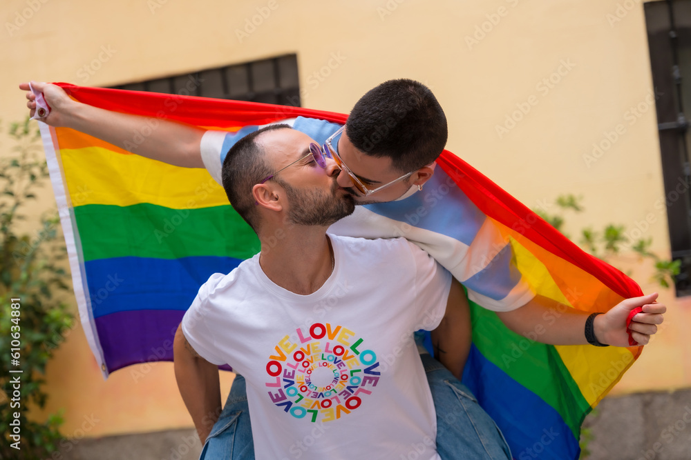 Homosexual male couple riding on top of back kissing each other at pride party with rainbow flag ...